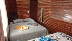 Quarto pequeno e estreito com parede de tijolos, cama com roupa de cama cinzenta e janela com moldura de metal.