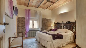 Quarto com parede de pedra, cama com roupa de cama branca, cabeceira de madeira e janela com cortinas.
