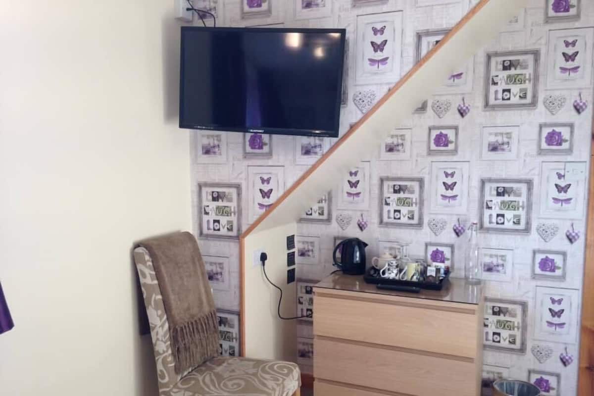 Quarto com televisão na parede, uma cadeira estampada, penteadeira de madeira e lixeira.
