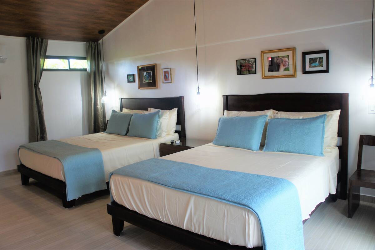 Quarto de hotel com duas camas, cada uma com colcha azul e branca, cabeceira de madeira e mesa de cabeceira com abajur.