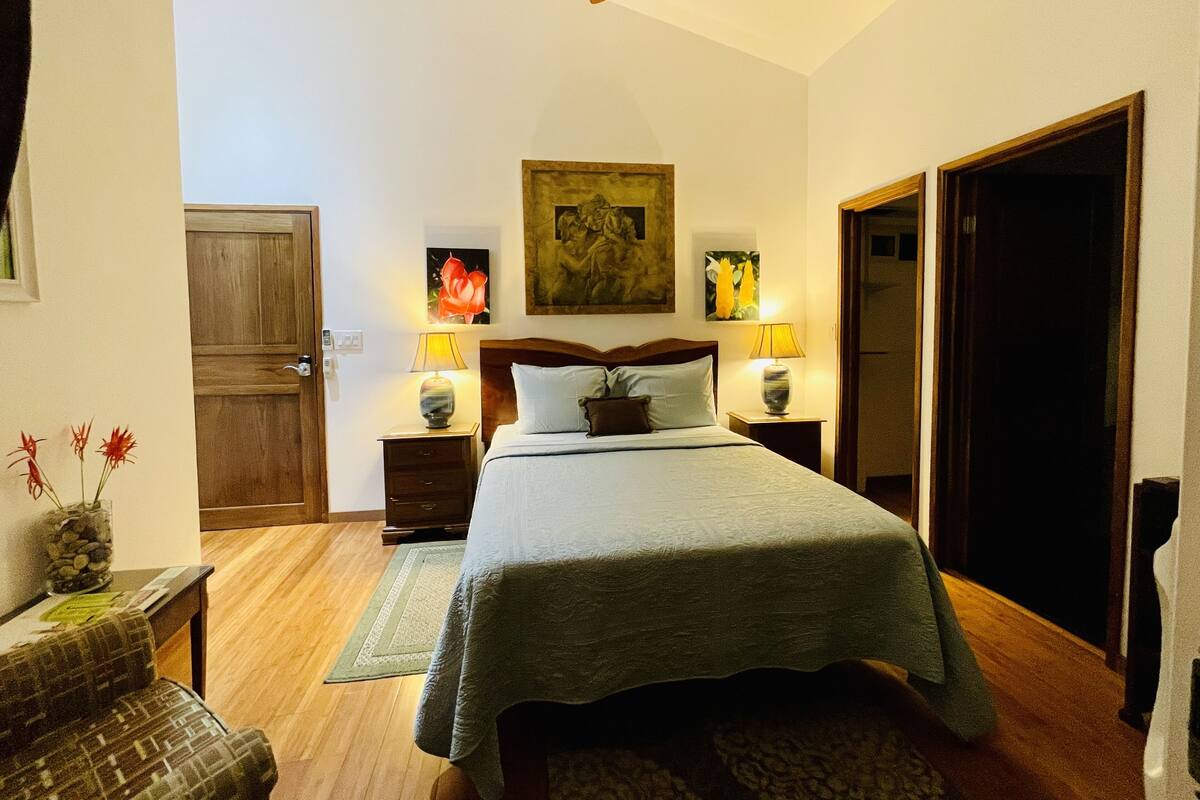 Um quarto com uma cama, duas mesas de cabeceira com luminárias, uma porta de madeira, um quadro na parede e uma pequena mesa com um vaso de flores.