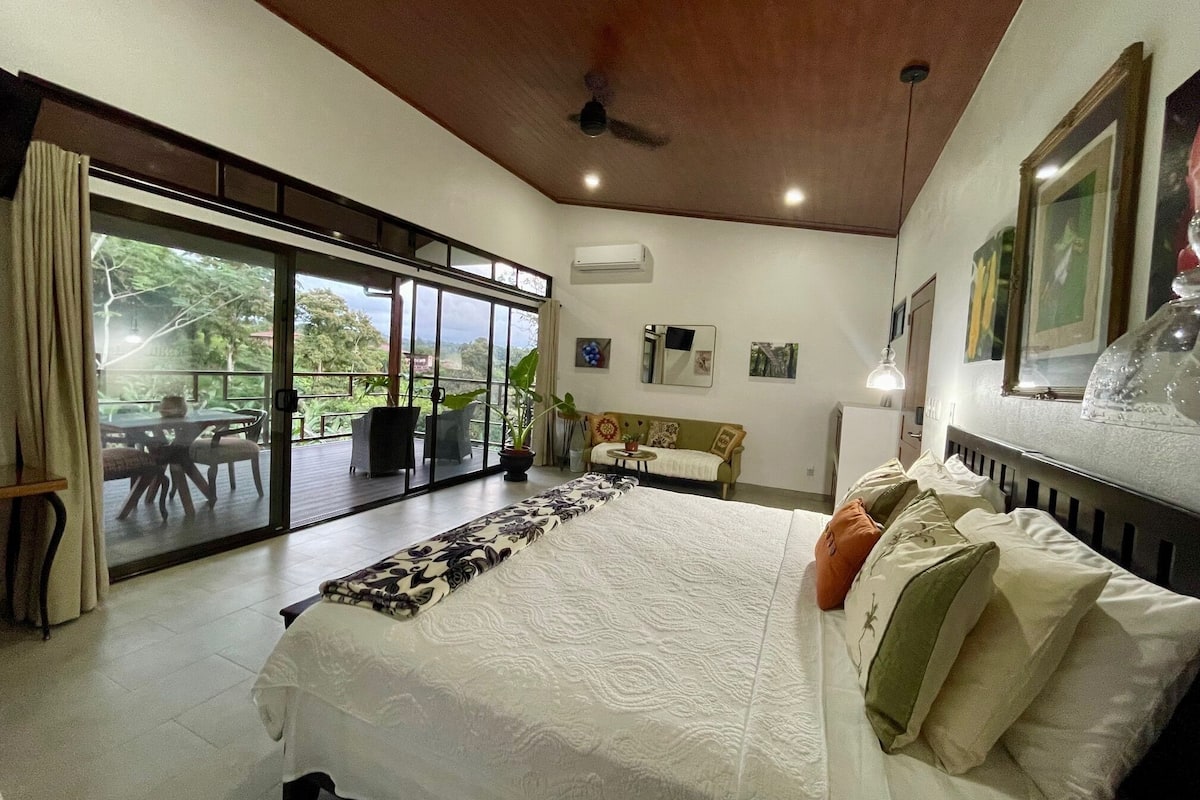 Quarto com uma cama grande, uma porta de correr de vidro que dá para uma área externa com mesa e cadeiras, e um ventilador de teto.