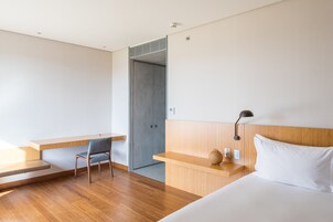 Quarto de hotel moderno com escrivaninha de madeira, cadeira, cama com roupa branca, mesa de cabeceira e luminária na parede.