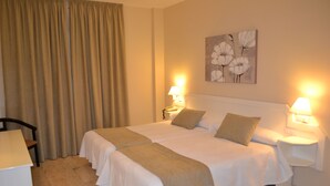 Uma cama de casal com duas almofadas, uma mesinha de cabeceira com abajur, um quadro na parede e uma cadeira.
