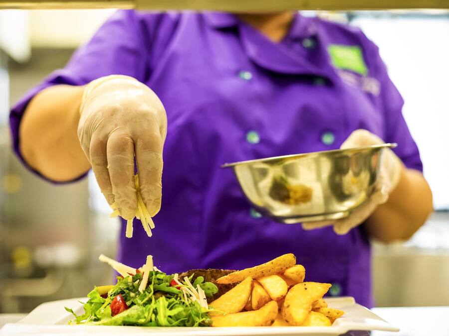 Uma pessoa vestida com uniforme e luvas roxas preparando comida, com um prato de batatas fritas e salada em primeiro plano.
