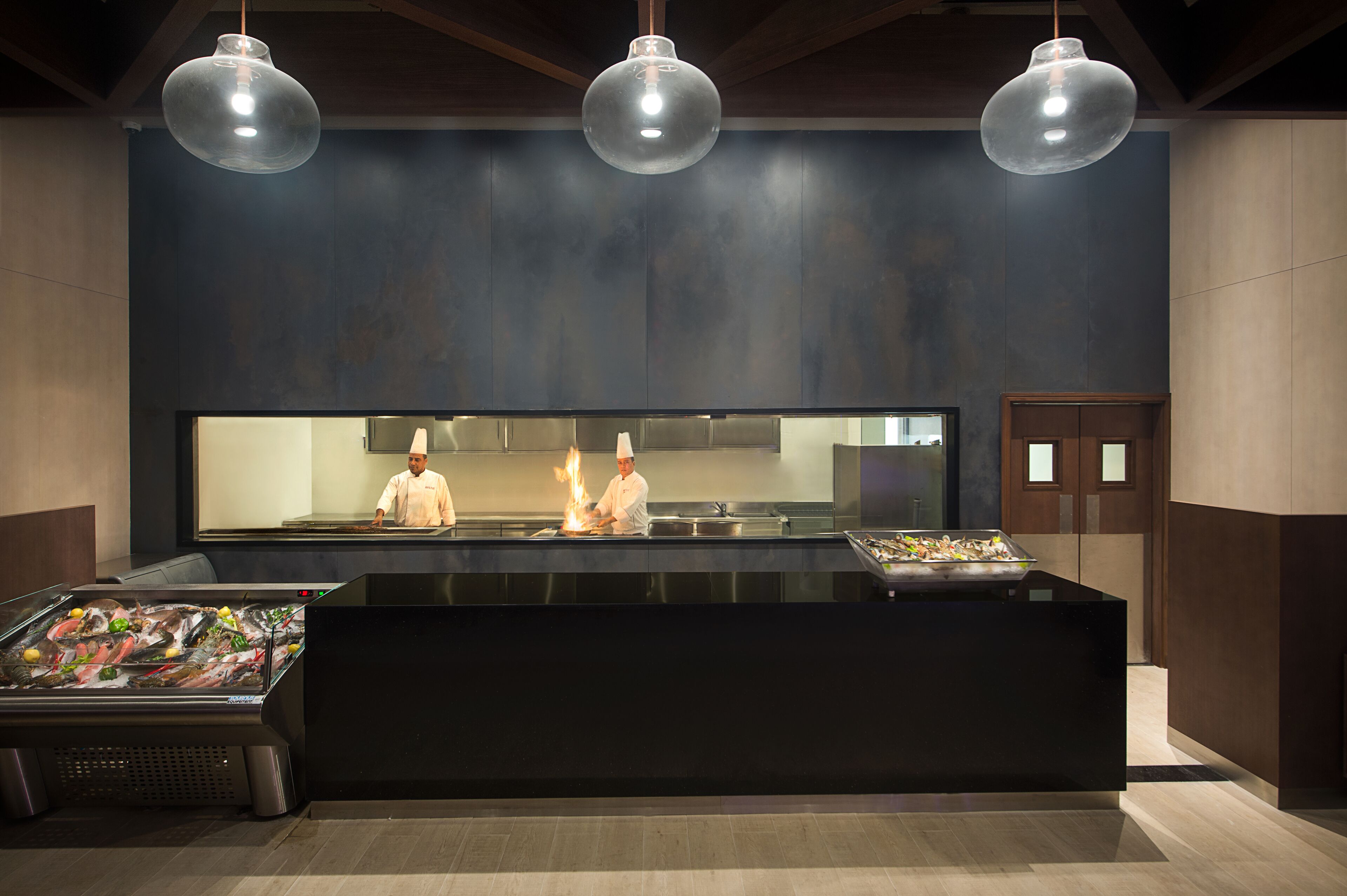 Cozinha moderna com chefs preparando comida, balcão preto grande e três luminárias pendentes.