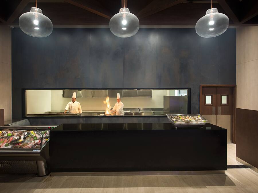 Cozinha moderna com chefs preparando comida, balcão preto grande e três luminárias pendentes.
