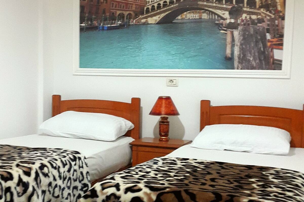 Quarto de hotel com duas camas, uma mesa de cabeceira com abajur e uma pintura grande de uma ponte sobre a água.