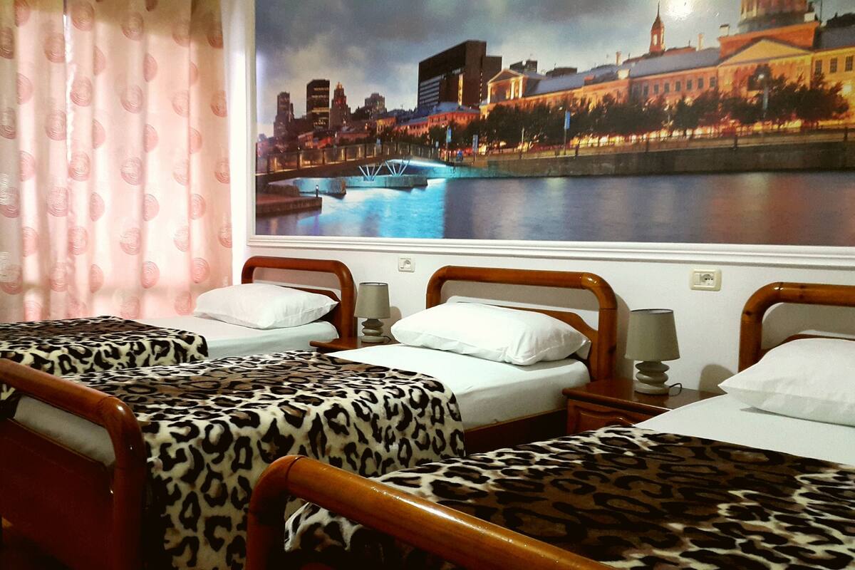 Quarto de hotel com duas camas, cada uma com colcha de padrão de leopardo, uma janela grande com vista para a cidade e cortina com padrão floral.