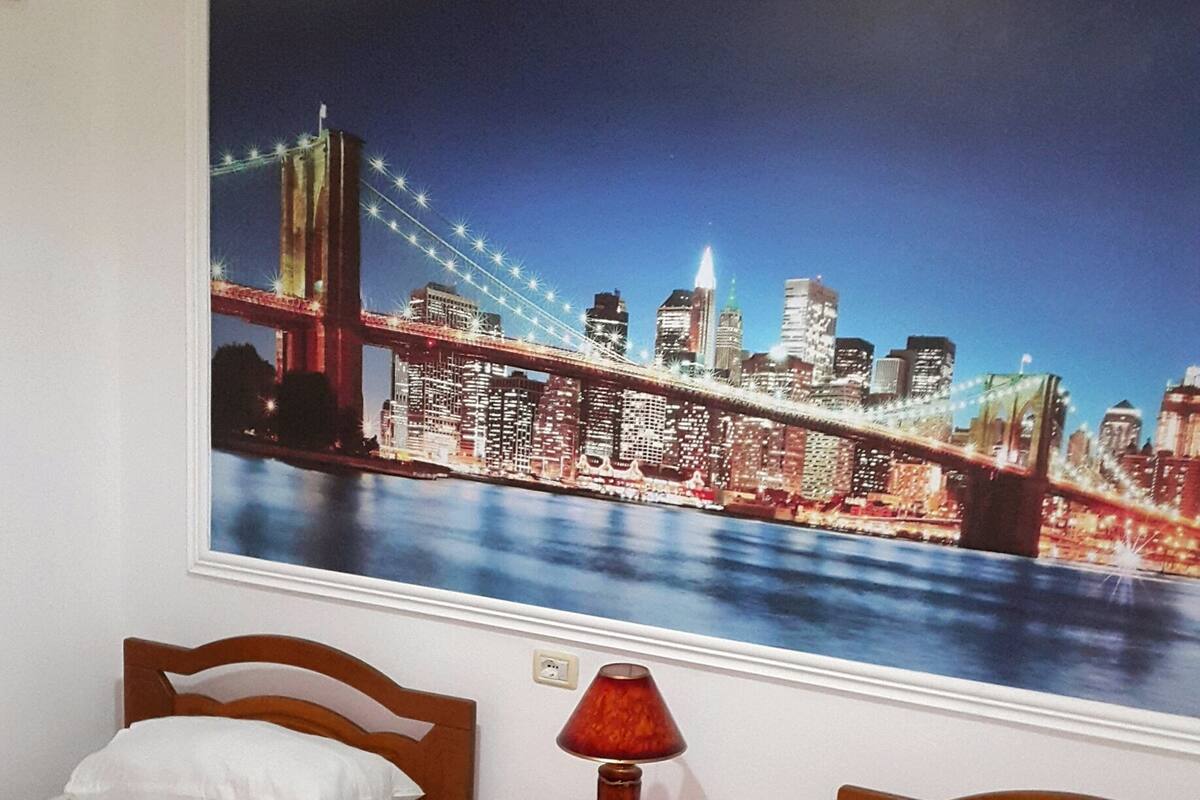 Quarto de hotel com duas camas, uma mesa de cabeceira, uma luminária e um grande mural com vista para a cidade.