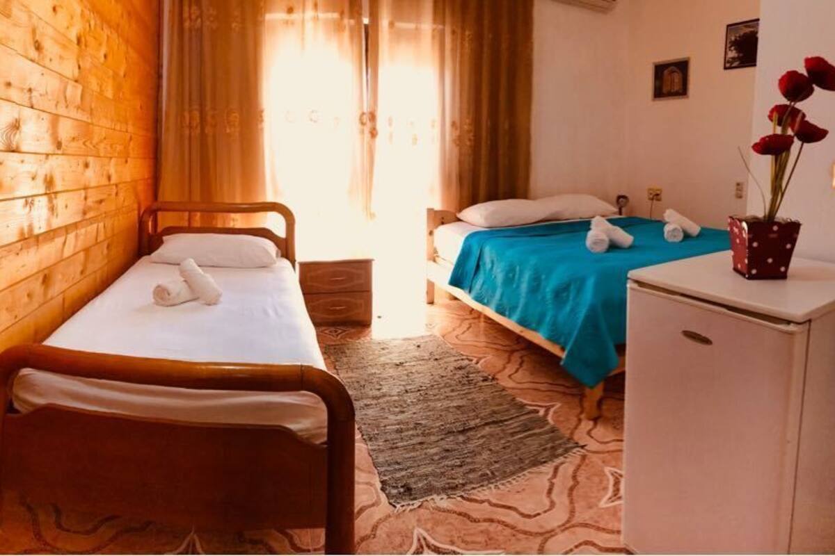 Quarto com duas camas de madeira, um pequeno armário branco e uma janela com cortinas.