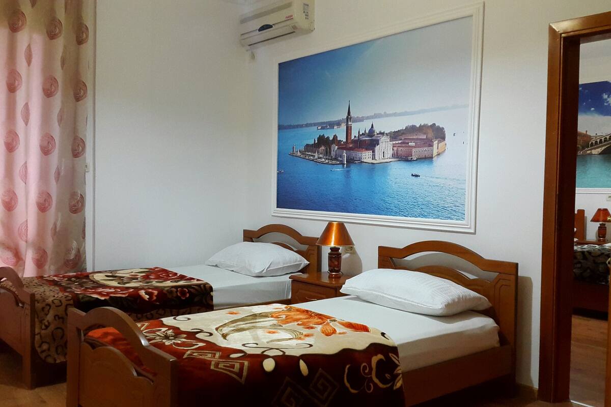 Quarto de hotel com duas camas de solteiro, uma mesinha de cabeceira com abajur, um quadro de uma cidade costeira e uma porta de madeira.