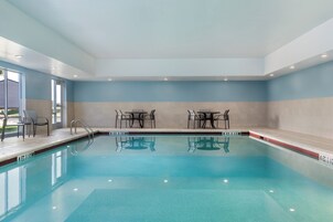 Piscina interna com água cristalina, cercada por paredes pintadas em tons de azul claro e bege, e equipada com mesas e cadeiras ao longo da borda.