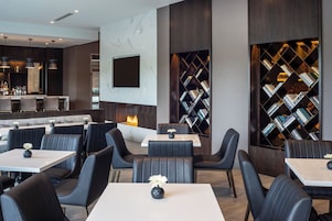 Interior de restaurante moderno com cadeiras de couro preto, mesas brancas, área de bar e lareira.