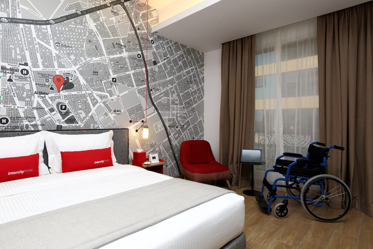 Quarto de hotel com uma cama grande, uma cadeira vermelha, uma cadeira de rodas e um mapa na parede.
