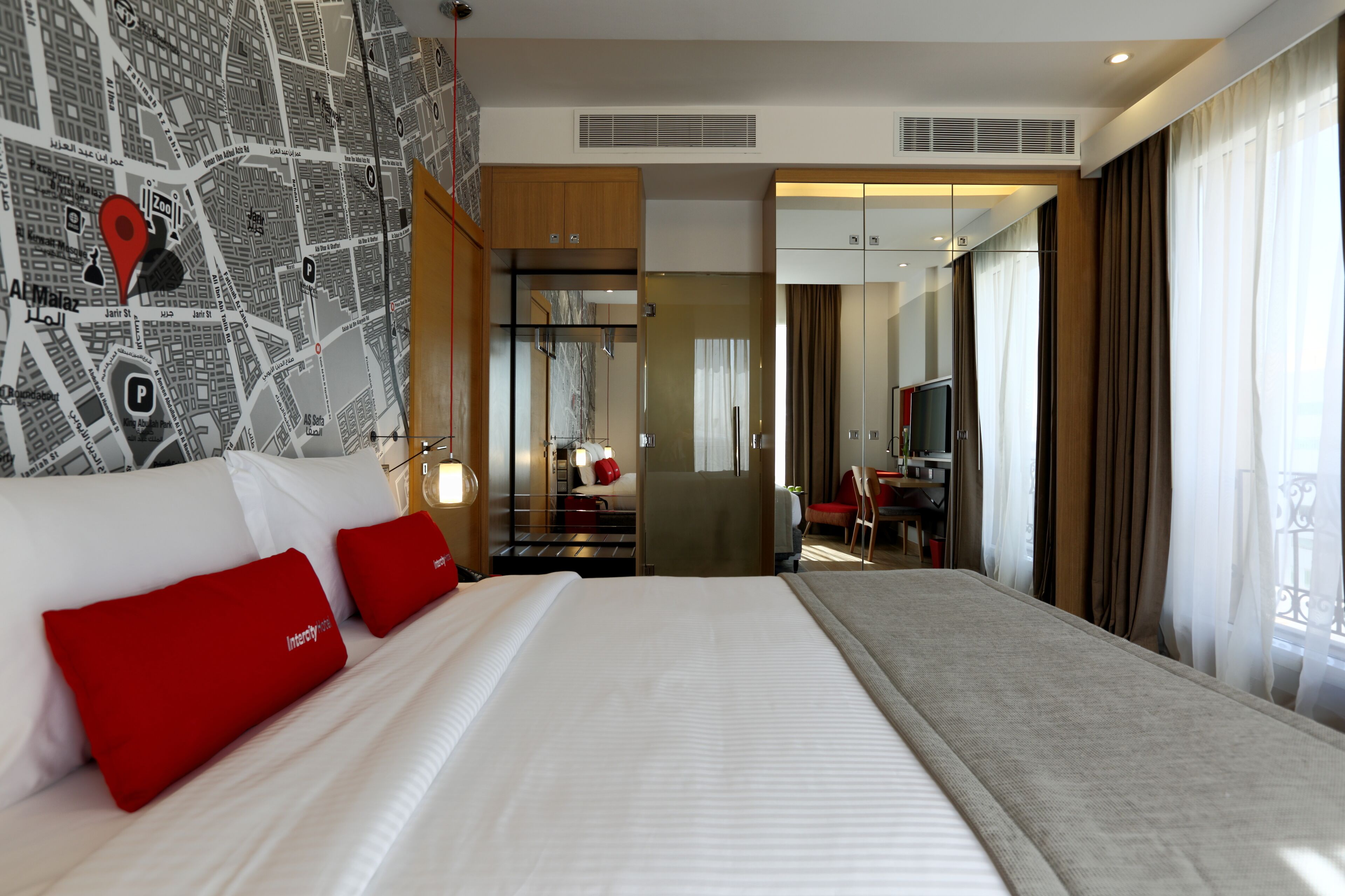 Quarto de hotel com uma cama grande, almofadas vermelhas, um mapa da cidade na parede, uma televisão e vista para a cidade pela janela.
