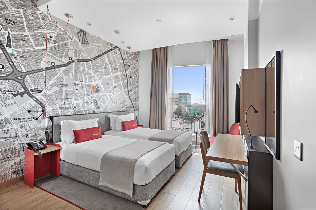 Quarto de hotel com duas camas, uma escrivaninha, uma cadeira, um mapa na parede e vista para a cidade.