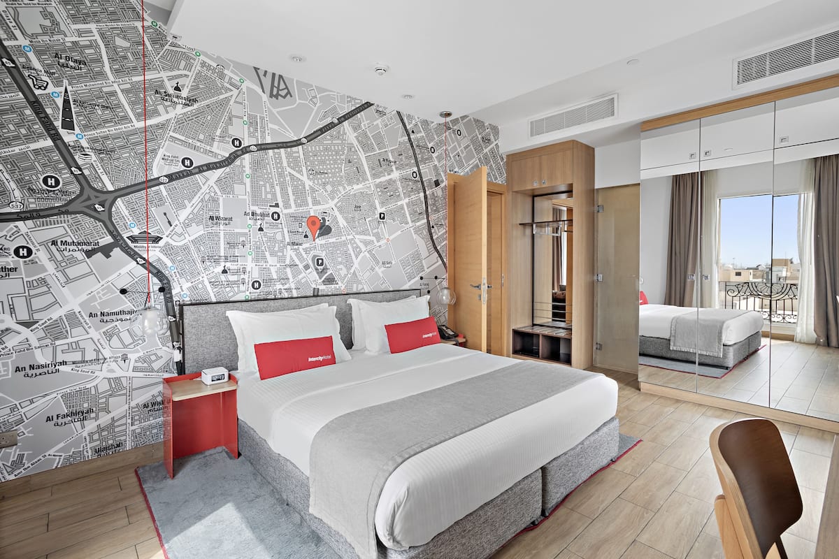 Quarto de hotel moderno com uma cama grande, mesa de cabeceira vermelha, parede com mapa, guarda-roupa com espelho e vista para a cidade pela janela.