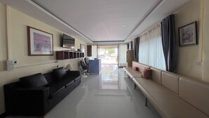 Um saguão de hotel com um sofá preto, um sofá bege, uma TV presa na parede e uma janela grande com cortinas.