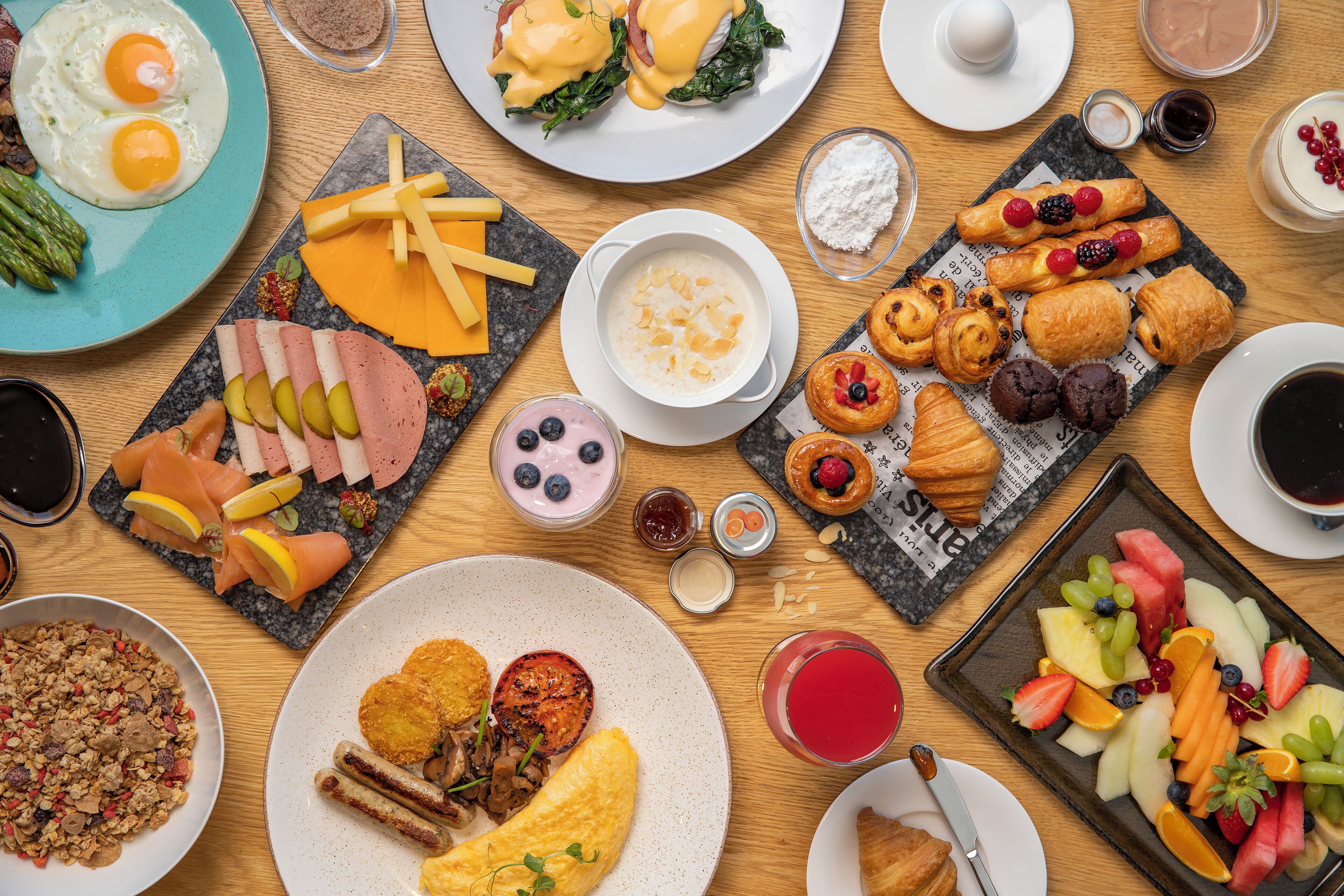 Um buffet de café da manhã com diversos pratos, incluindo ovos, presunto, queijo, frutas, pães e bebidas.