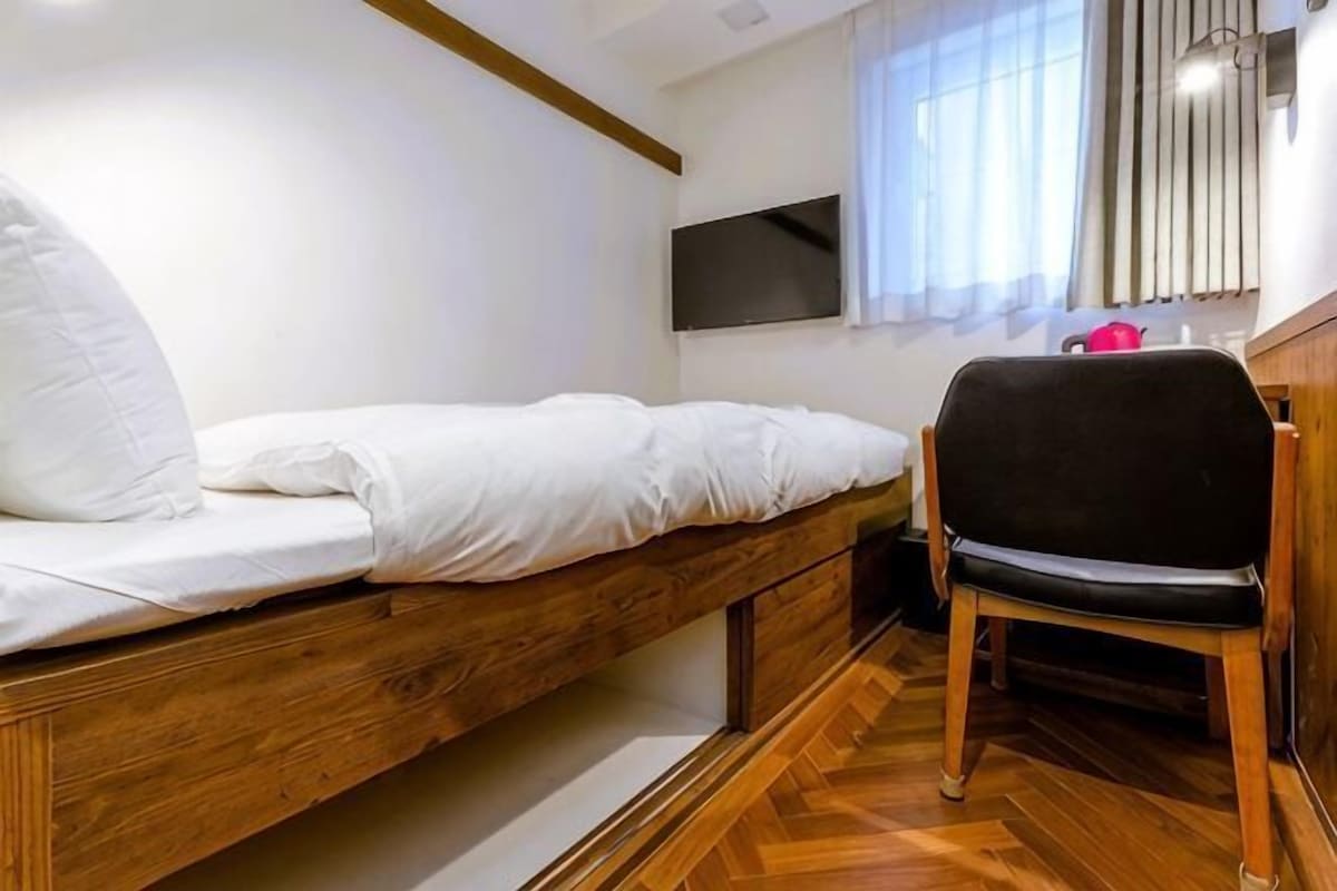 Quarto com cama, poltrona, televisão e janela com persianas.