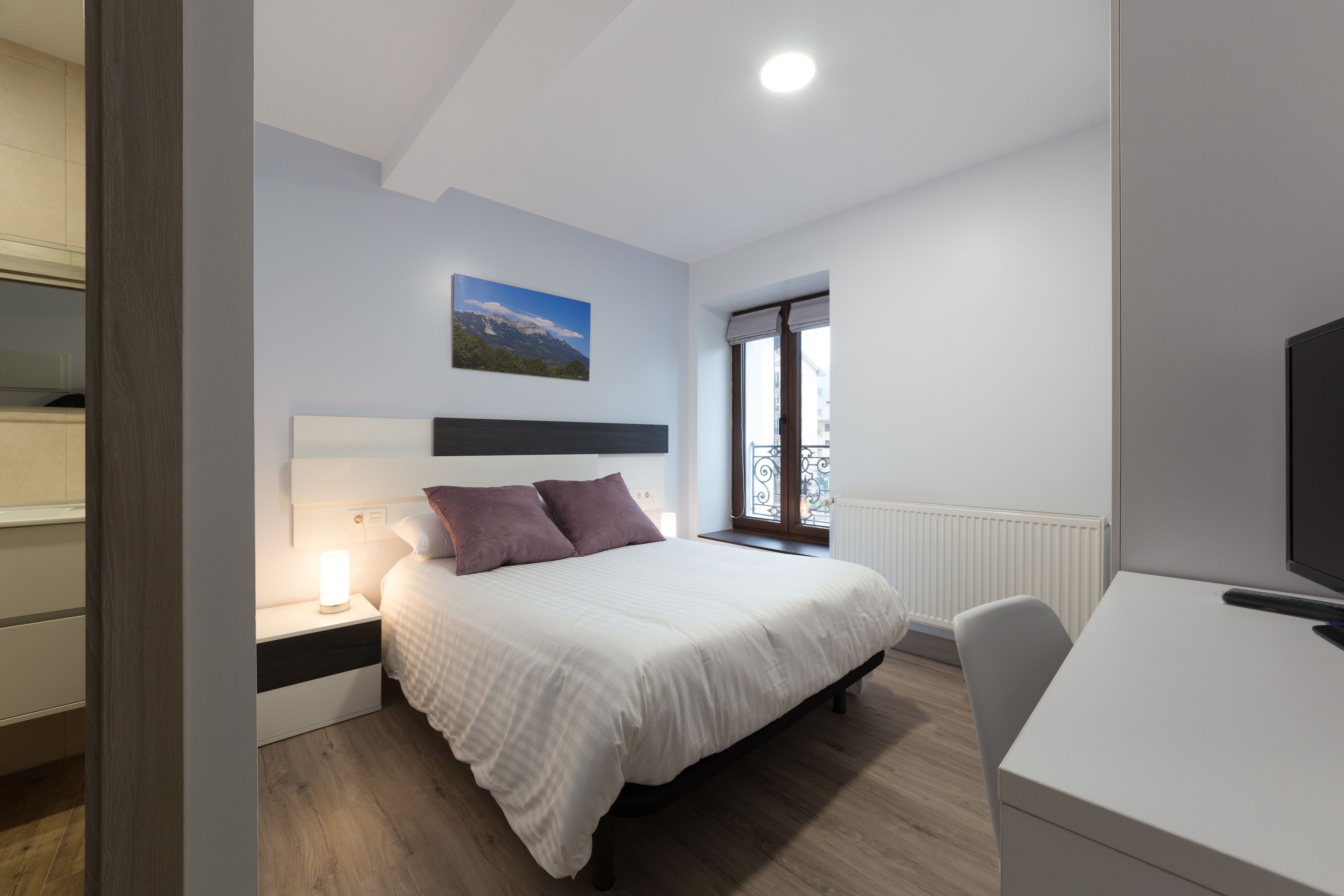Quarto com cama, mesa de cabeceira, abajur e escrivaninha com cadeira.
