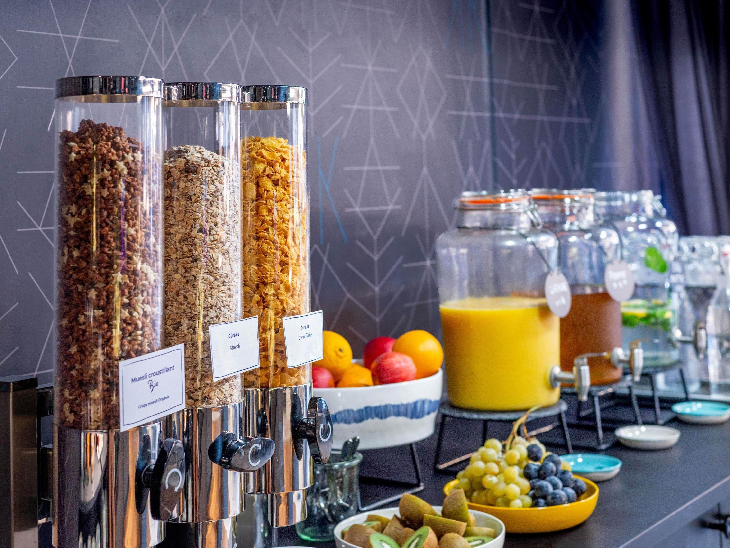 Buffet de café da manhã com recipientes identificados de granola, muesli e flocos de milho, uma cesta de frutas e um dispensador de suco.