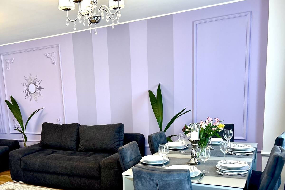 Uma sala de estar moderna com área de jantar, apresentando um sofá, mesa de jantar posta com pratos e copos, e um lustre.
