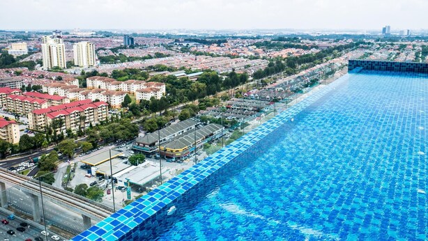 Uma piscina grande com um deck ladrilhado, cercada por edifícios residenciais e uma rodovia.