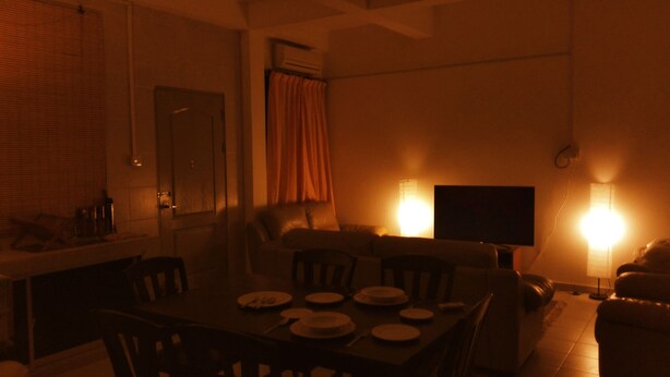 Uma sala de estar com pouca luz, com uma mesa de jantar posta para quatro pessoas, um sofá e uma televisão.