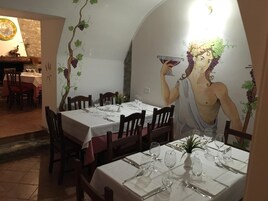 Restaurante com toalhas de mesa brancas, cadeiras de madeira e um mural de um homem segurando uma taça de vinho.