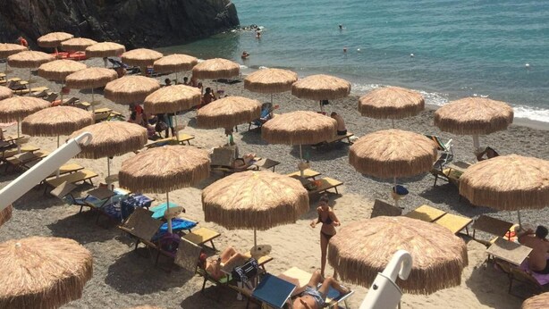 Uma praia com guarda-sóis de palha e espreguiçadeiras, falésias rochosas e mar calmo.
