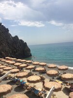 Uma praia com guarda-sóis de palha e espreguiçadeiras, falésias rochosas e mar calmo.