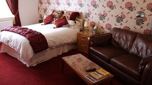 Quarto de hotel com cama, sofá de couro marrom, mesa de cabeceira de madeira e parede com padrão floral.