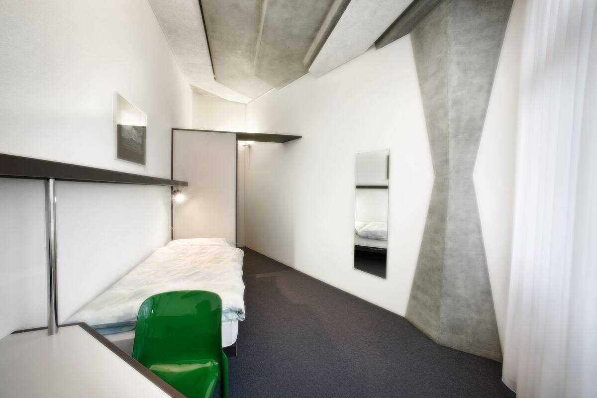 Quarto de hotel moderno com uma cama de solteiro, uma escrivaninha, uma cadeira, um espelho e uma janela com cortinas.
