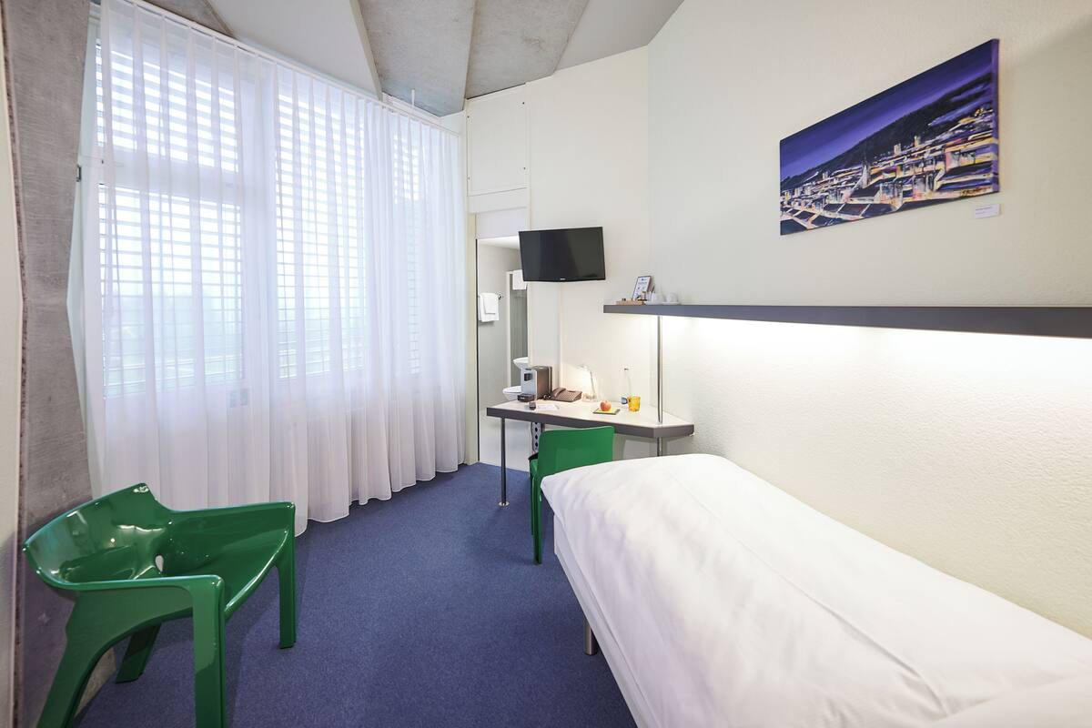 Quarto de hotel com uma cama, uma escrivaninha com cadeira, uma televisão na parede e uma janela com cortinas.