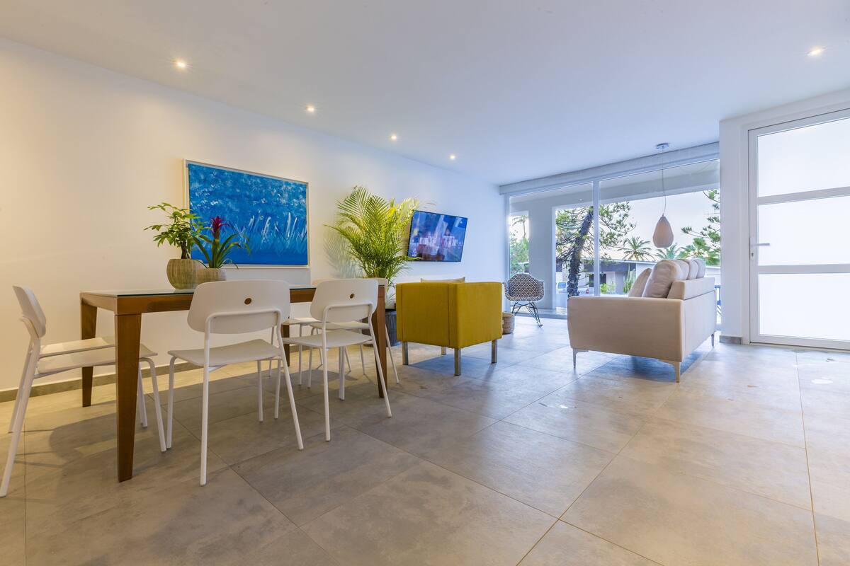 Uma sala de estar moderna com área de jantar, apresentando um sofá, cadeiras, uma mesa e uma grande janela com vista para o verde.