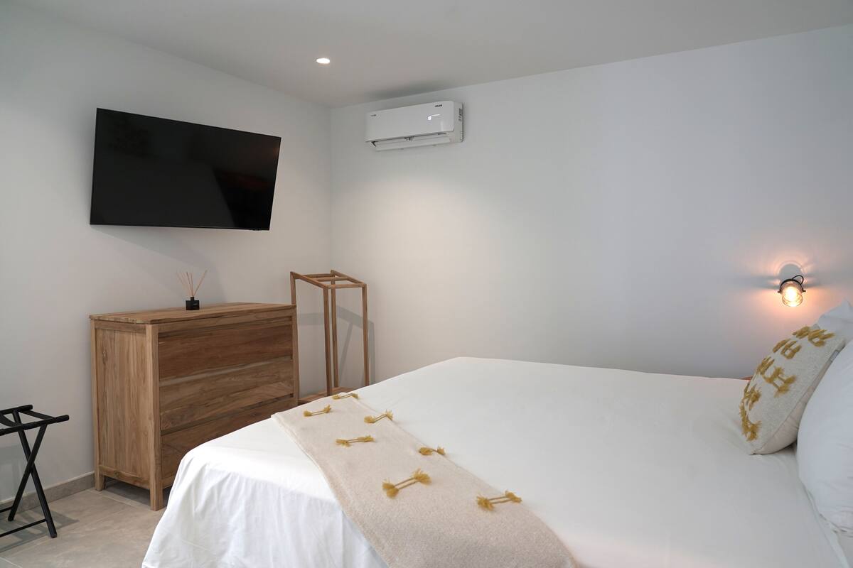 Quarto com cama, televisão na parede, cómoda de madeira e luminária fixada na parede.