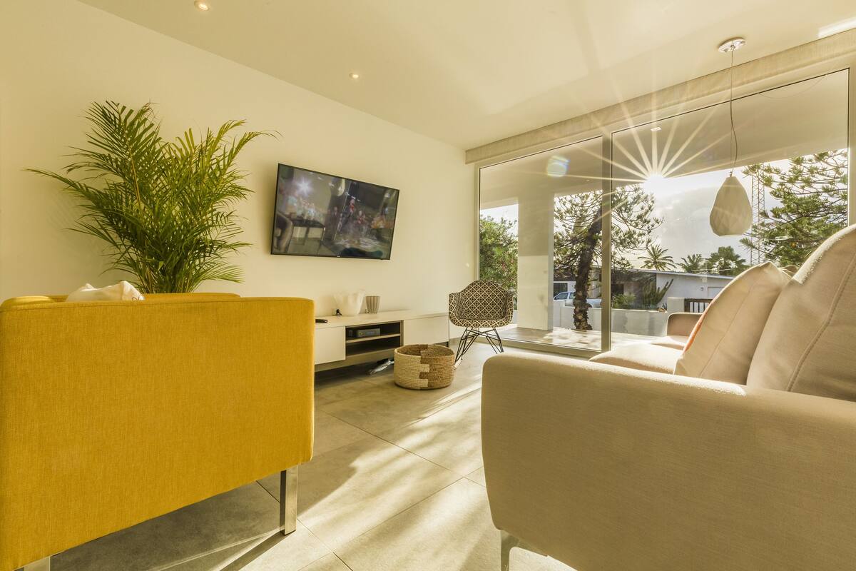 Uma sala de estar moderna com um sofá, uma poltrona amarela, uma planta grande e uma televisão na parede.