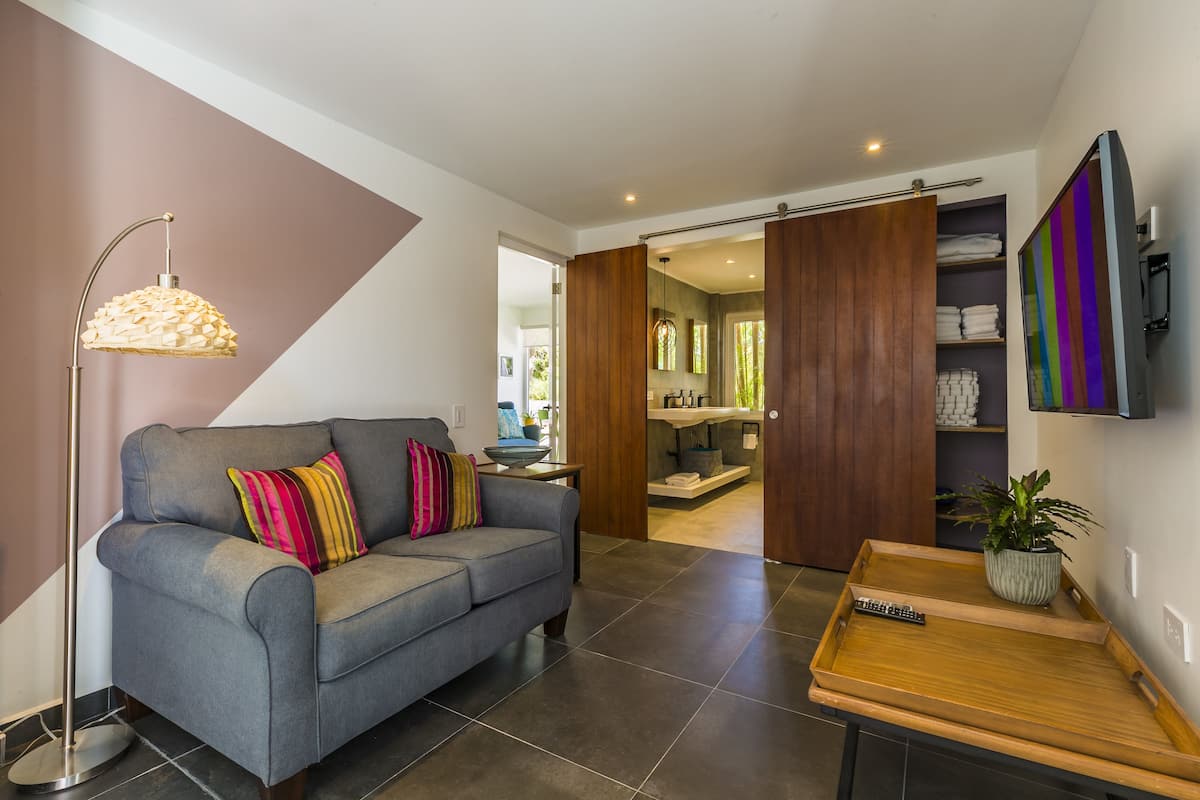 Uma sala de estar moderna com um sofá cinza, almofadas coloridas, um abajur, uma mesa lateral de madeira e uma TV fixada na parede.