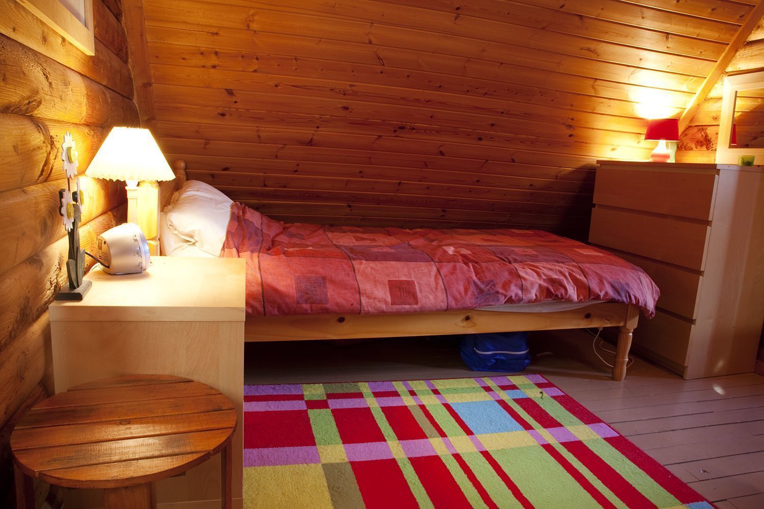 Um quarto pequeno e acolhedor com uma cama de madeira beliche, uma escrivaninha com abajur e um tapete colorido.