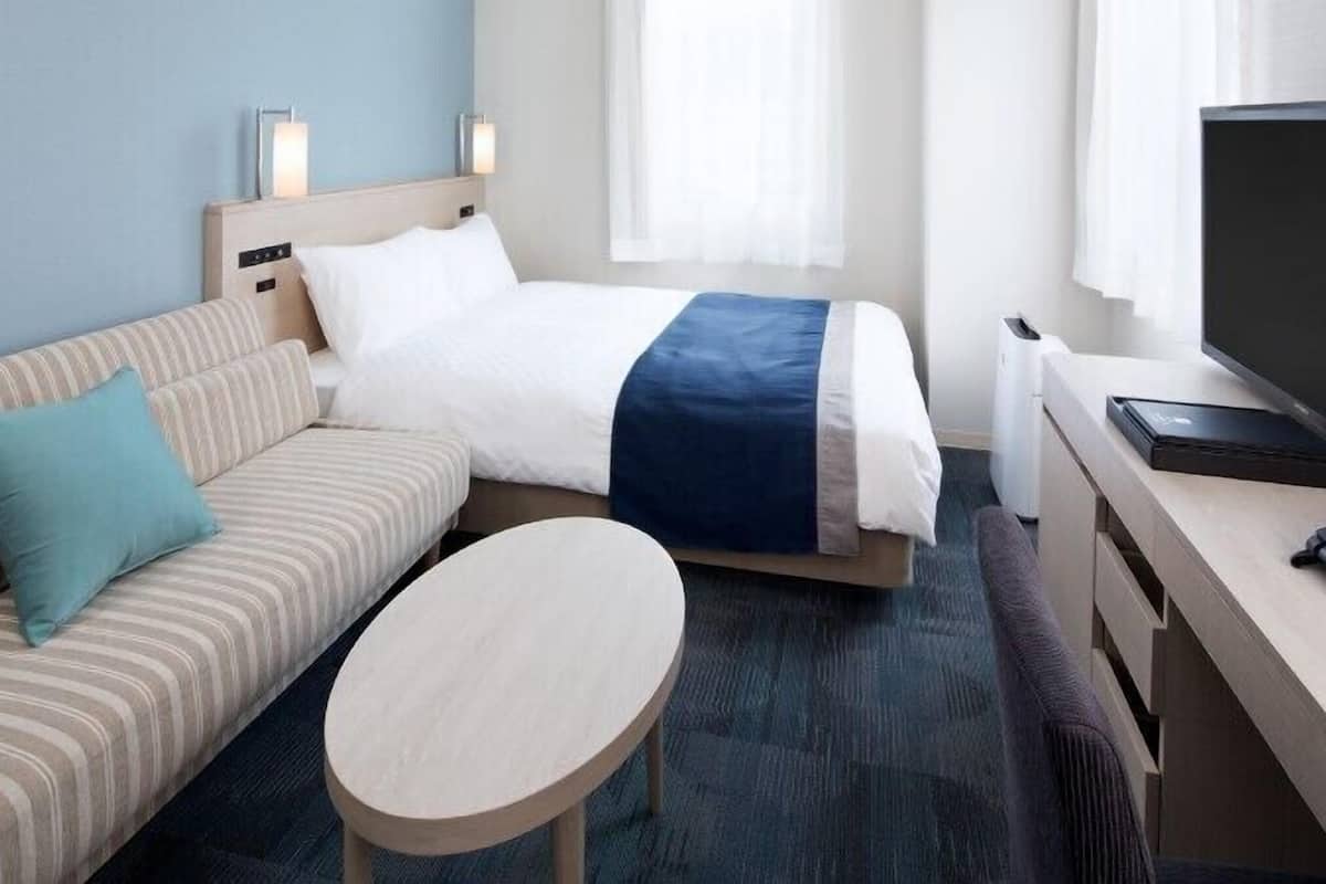 Quarto de hotel com uma cama, um sofá, uma mesinha, uma escrivaninha com um computador e uma televisão.