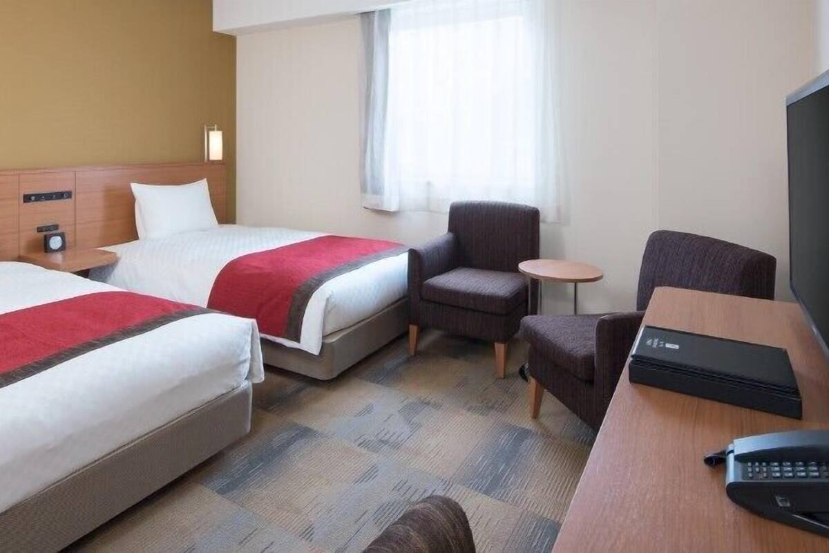 Quarto de hotel com duas camas, uma escrivaninha com telefone, uma cadeira, uma mesinha e uma televisão.