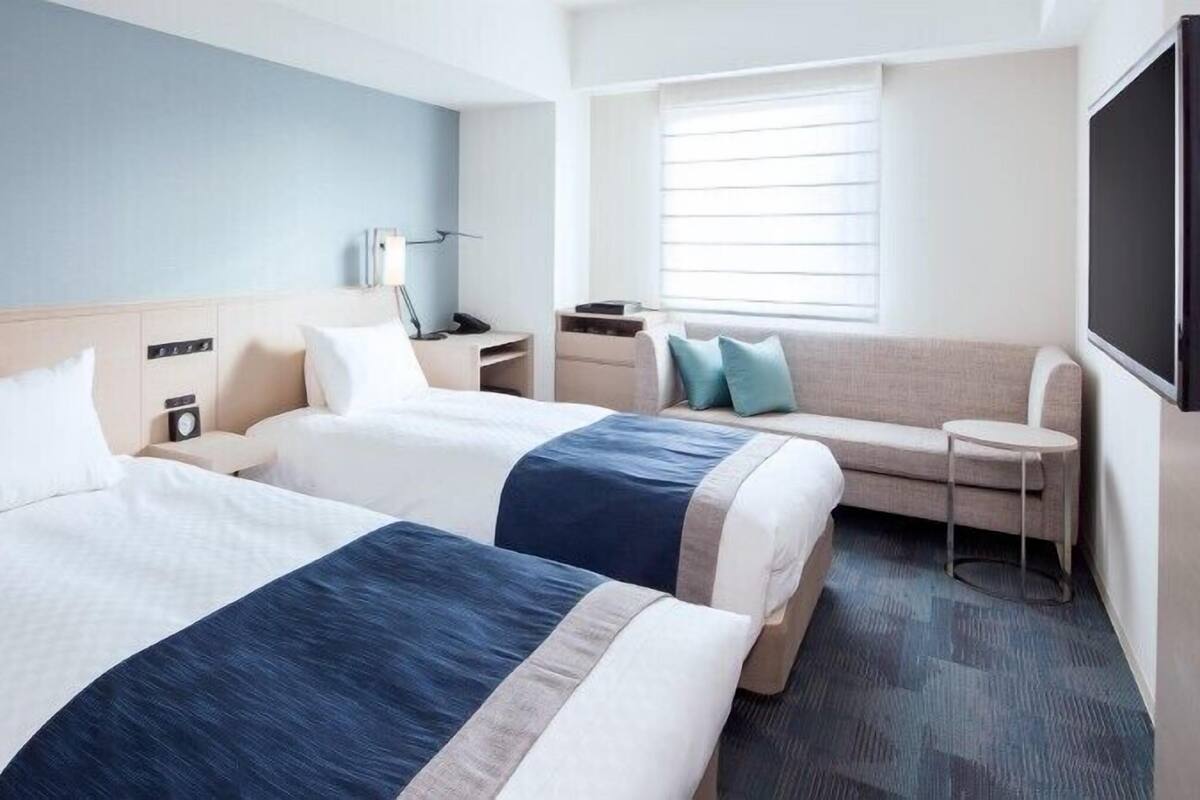 Quarto de hotel com duas camas, um sofá, uma televisão e uma janela com persianas.