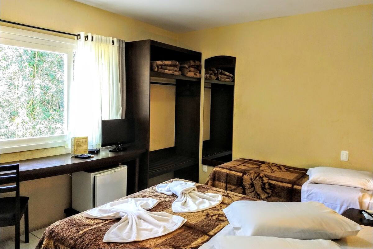 Quarto de hotel com cama, escrivaninha, computador e janela com cortinas.