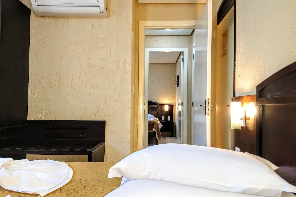 Quarto de hotel com cama, escrivaninha e cadeira. Uma porta dá acesso a outro espaço.
