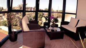 Uma varanda ensolarada com mobiliário de vime, plantas em vasos e vista para a paisagem urbana.