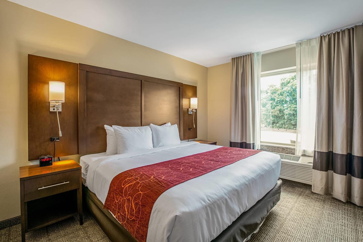 Quarto de hotel com uma cama grande, duas mesas de cabeceira, um criado-mudo, manta vermelha, cabeceira, duas luminárias de parede e uma janela com cortinas.
