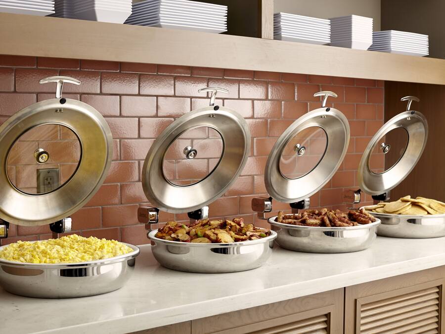 Disposição de buffet com quatro tigelas de aço inoxidável contendo pratos diversos, um fundo de parede de tijolos e uma prateleira de madeira acima.