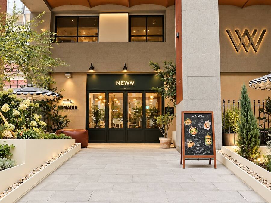 Entrada de hotel com fachada preta, placa com 'NEWW NOMADIA' e quadro de cardápio.